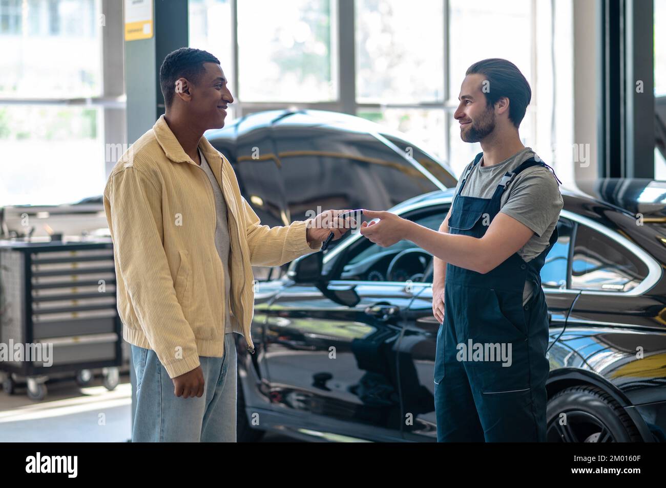Service. Dark-skinned smiling man holding out car key to bearded ...