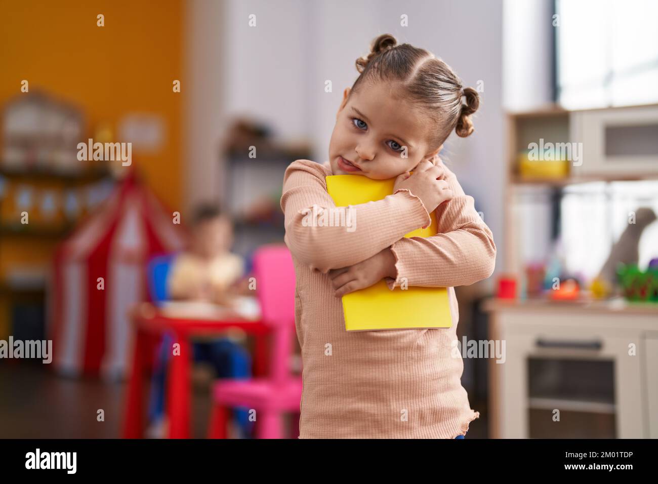 Adorable hispanic girl student hugging book standing at kindergarten ...