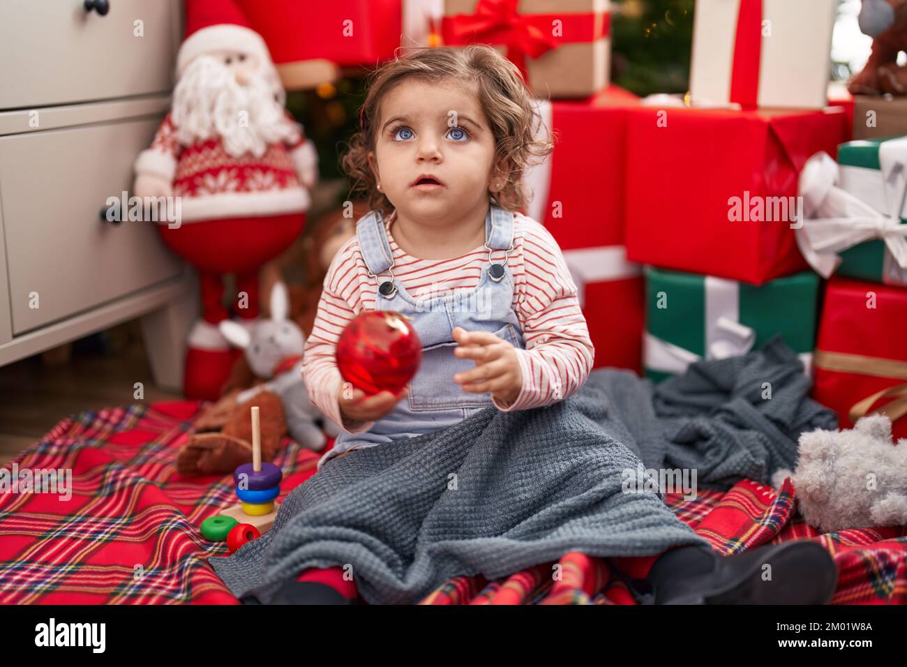 Adorable hispanic girl holding ball decoration sitting on floor by ...