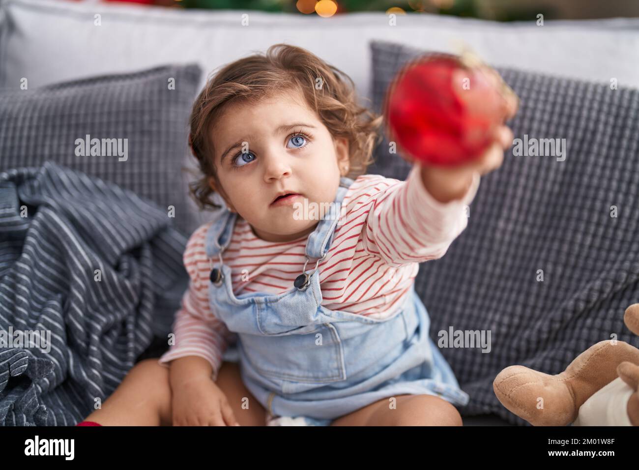 Adorable hispanic girl holding ball decoration sitting on sofa by ...