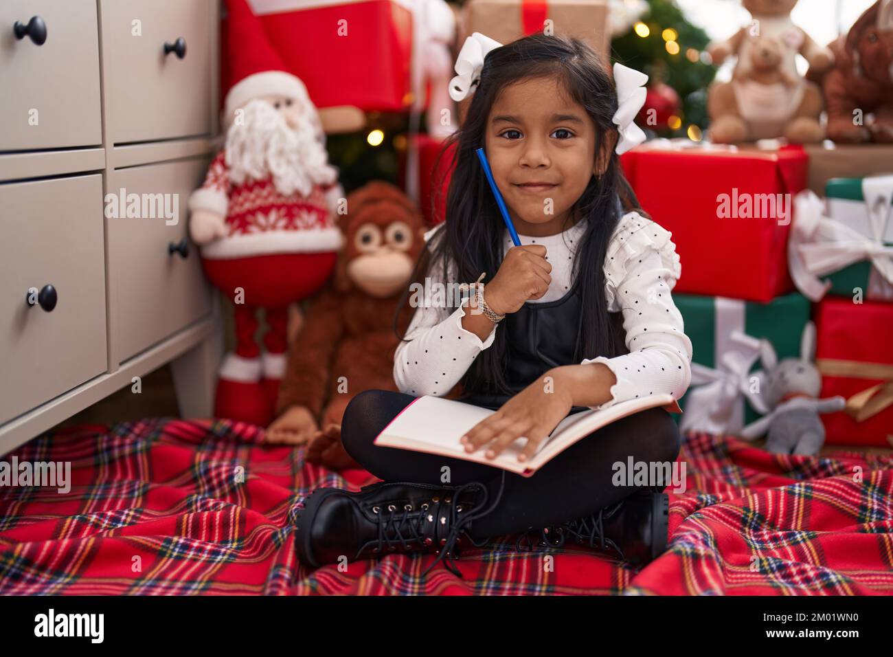 Adorable hispanic girl writing on notebook sitting on floor by ...
