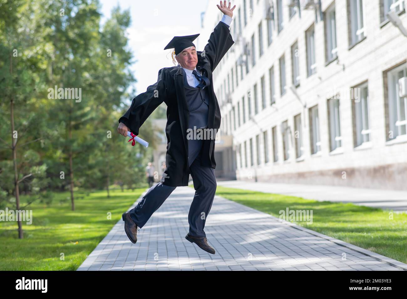 Old happy man in graduation gown jumping outdoors and holding diploma ...