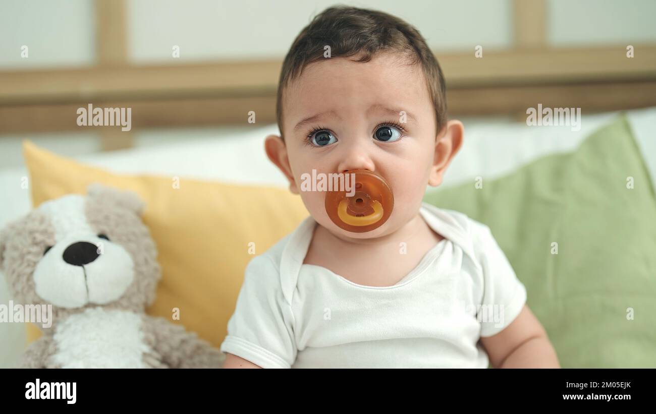 Adorable hispanic baby sucking pacifier sitting on bed at home Stock ...