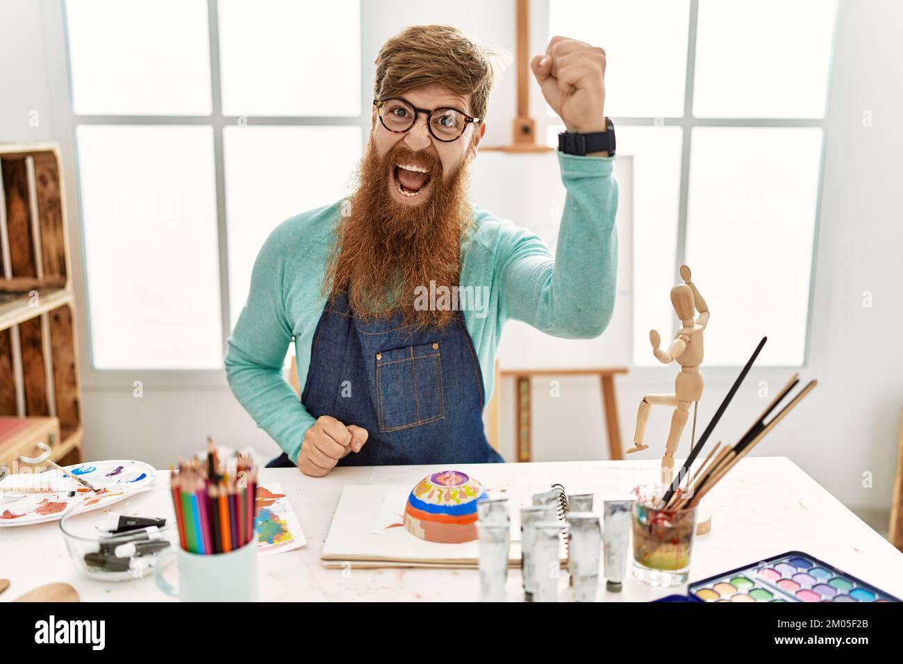 Redhead man with long beard painting clay bowl at art studio angry and ...