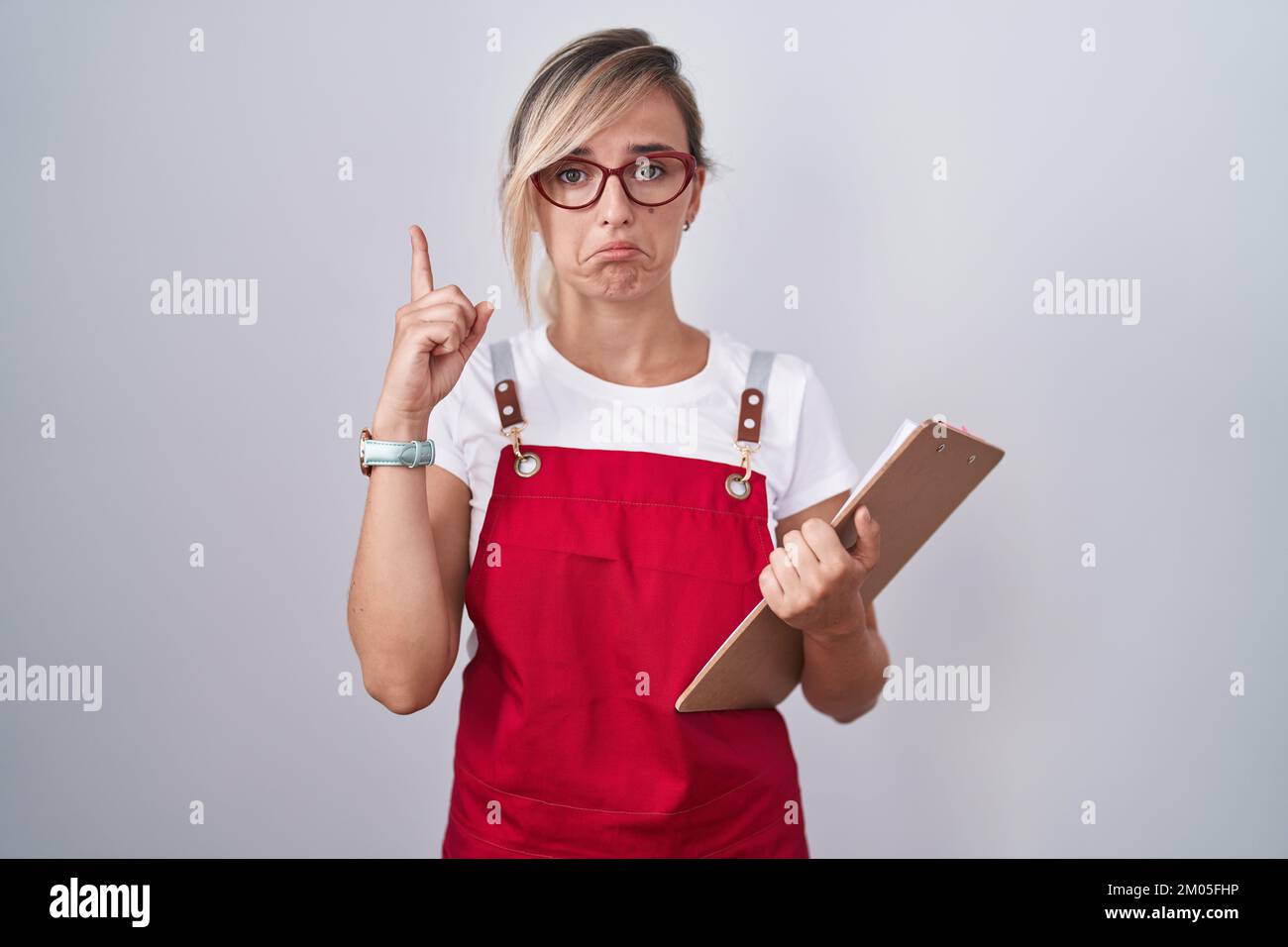Young blonde woman wearing waiter uniform holding clipboard pointing up ...