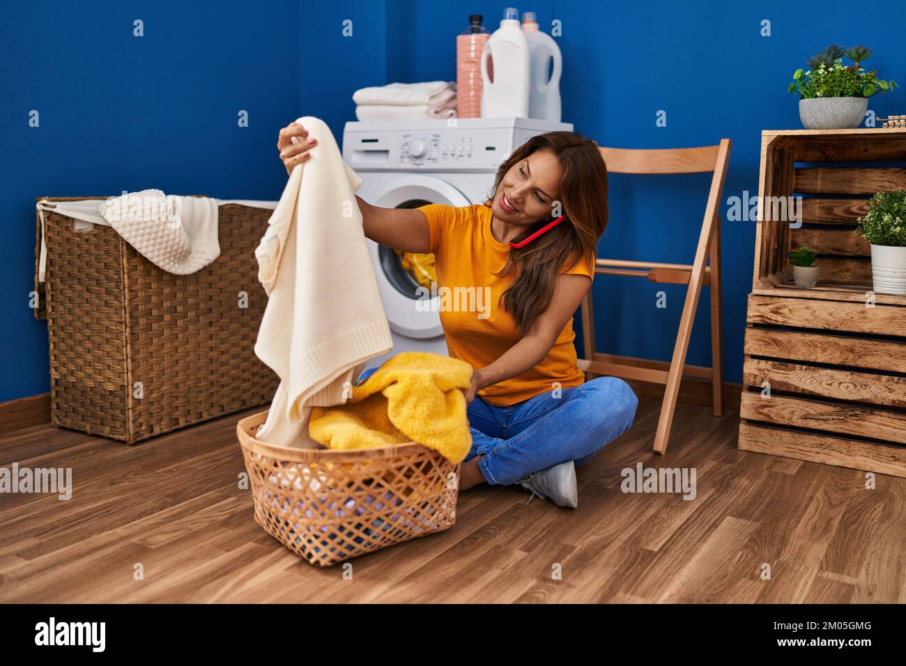 Young latin woman talking on the smartphone holding clothes at laundry ...