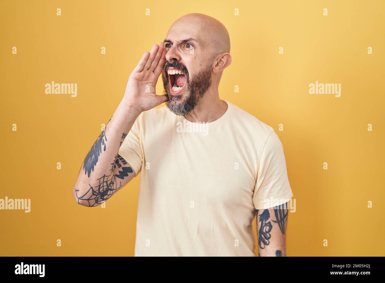 Hispanic man with tattoos standing over yellow background shouting and ...