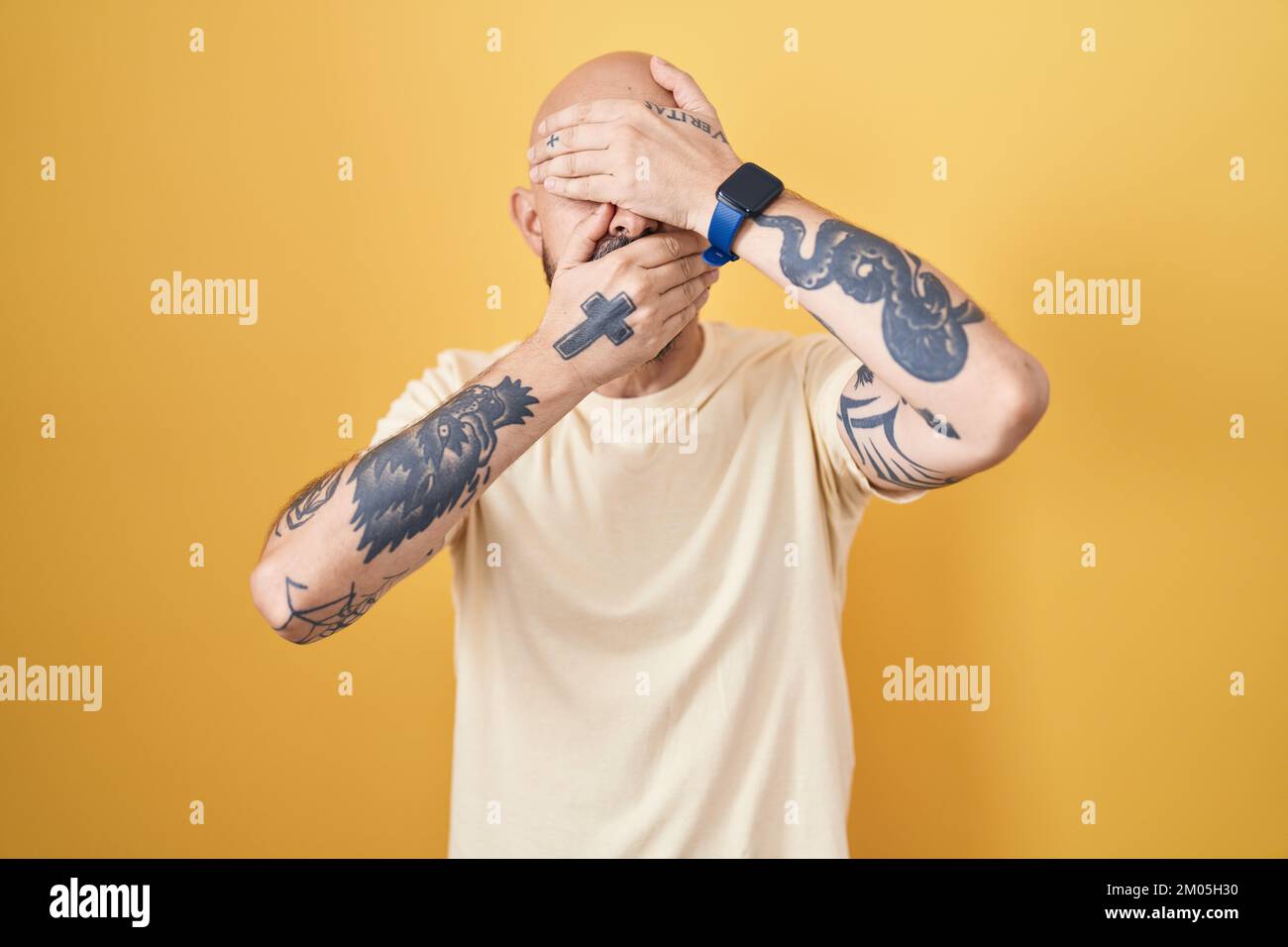 Hispanic man with tattoos standing over yellow background covering eyes ...