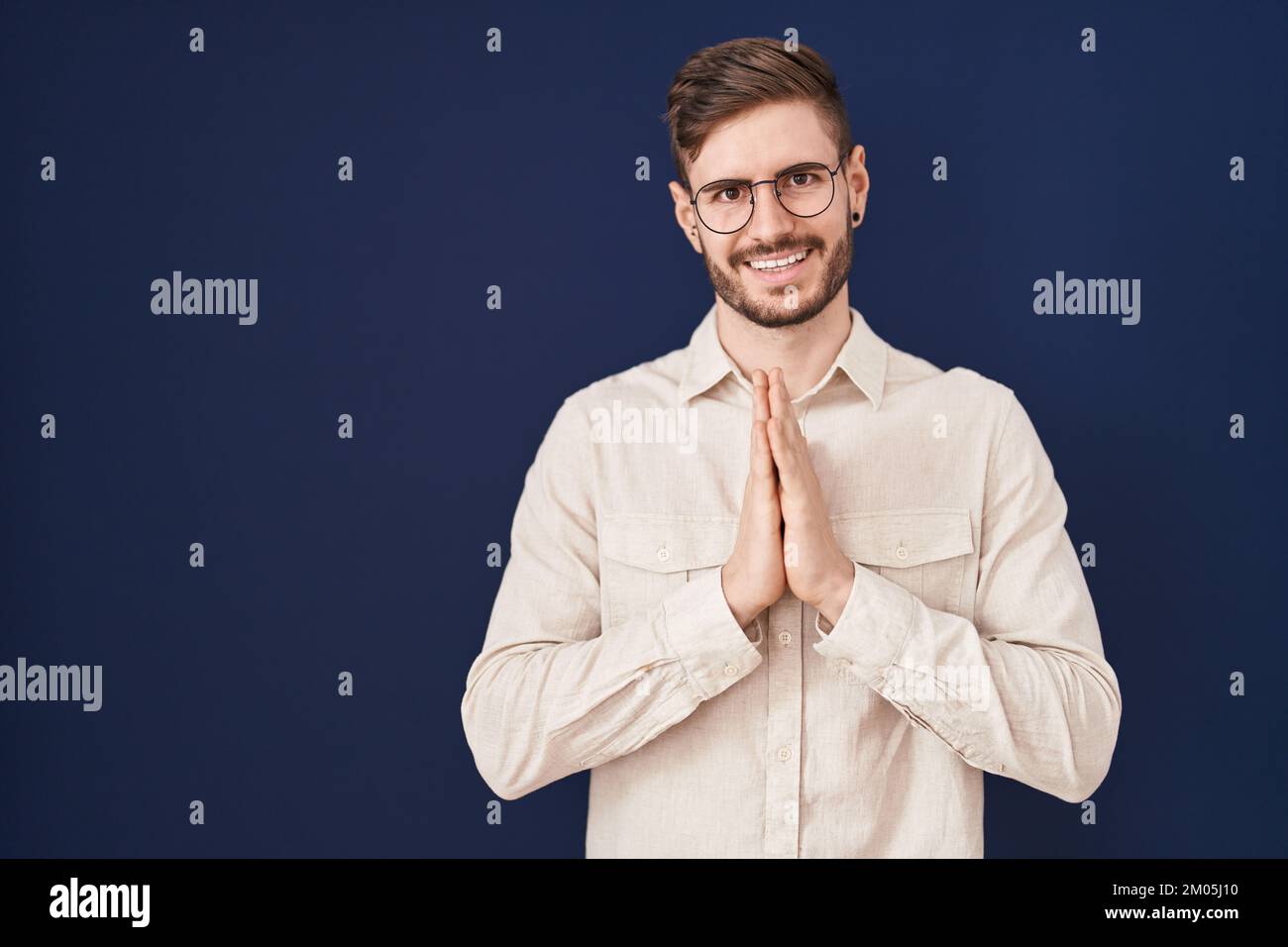 Hispanic man with beard standing over blue background praying with ...