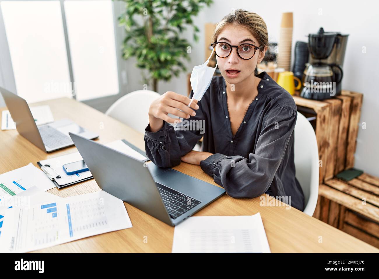 Young blonde woman working at the office wearing safety mask scared and ...