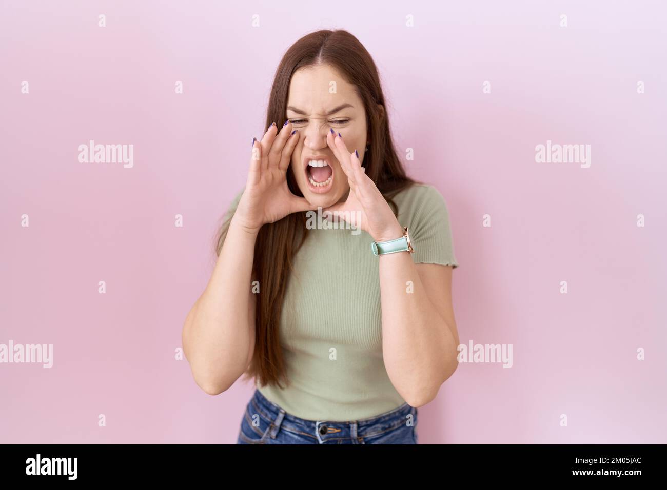 Beautiful brunette woman standing over pink background shouting angry ...