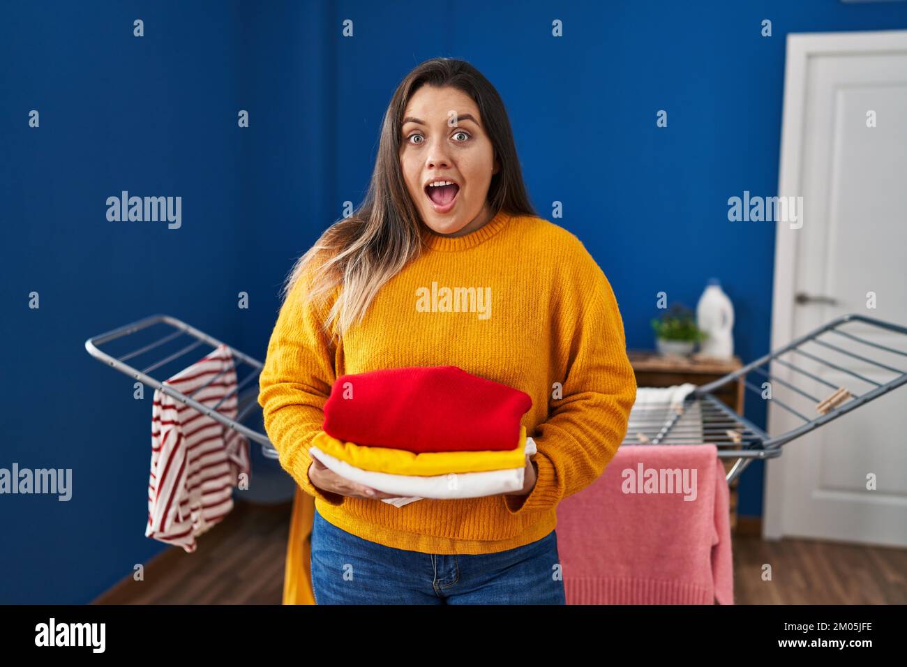 Young hispanic woman holding clean and folded laundry celebrating crazy and amazed for success ...