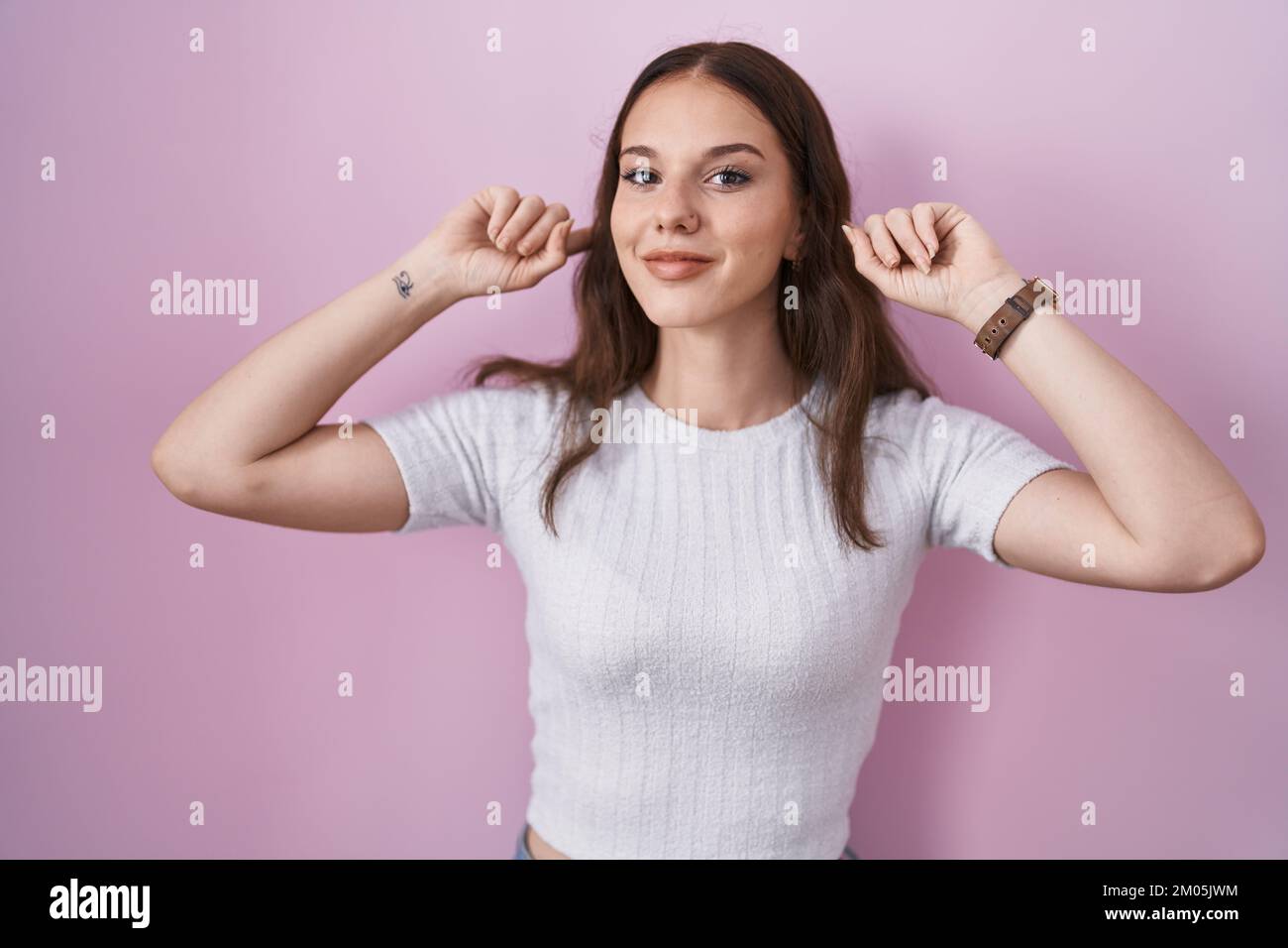 Young hispanic girl standing over pink background smiling pulling ears ...