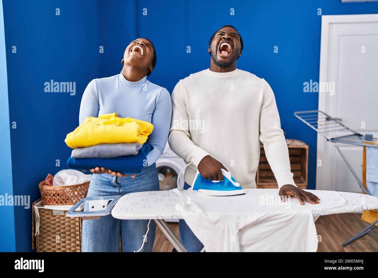 Young african american couple ironing clothes at laundry room angry and mad screaming frustrated ...