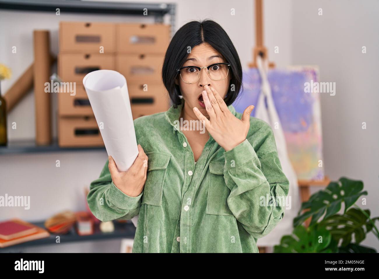 Young asian woman holding paper at art studio covering mouth with hand ...