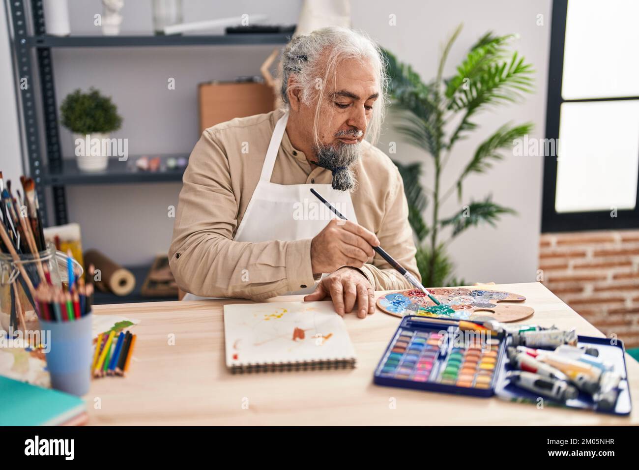 Middle age grey-haired man artist drawing on notebook with relaxed expression at art studio ...