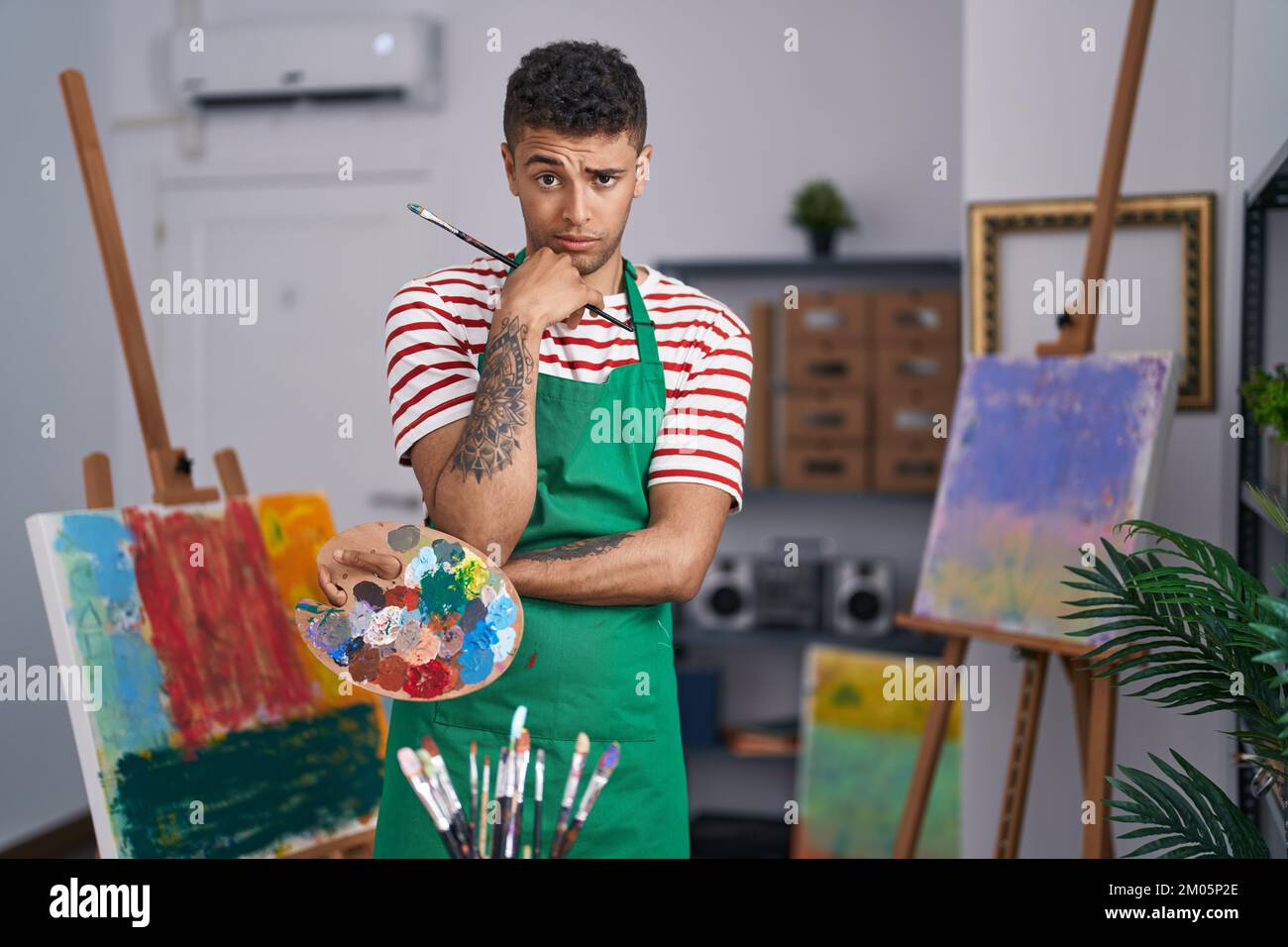 Brazilian young man holding painter palette at artist studio skeptic ...