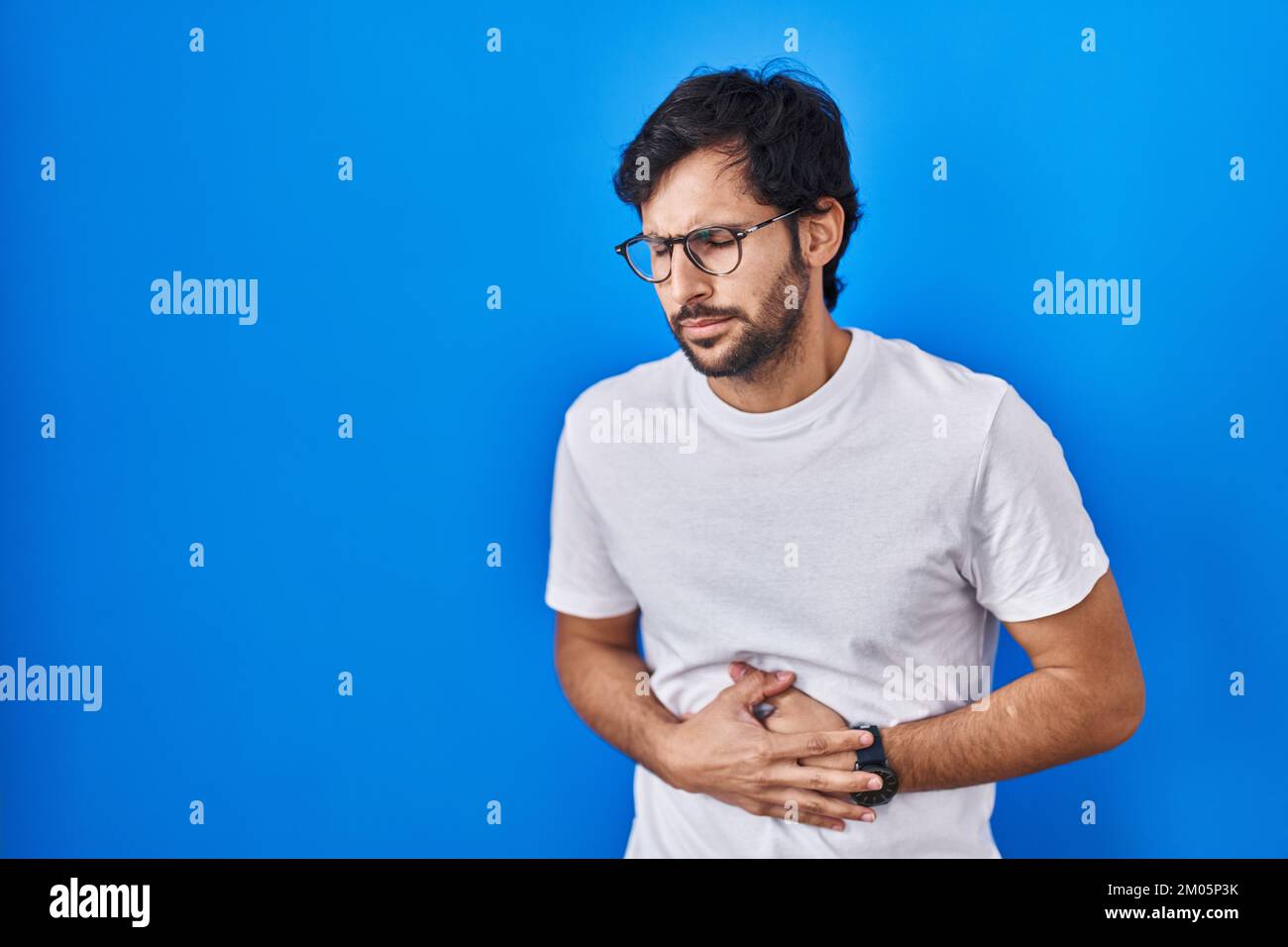 Handsome latin man standing over blue background with hand on stomach ...