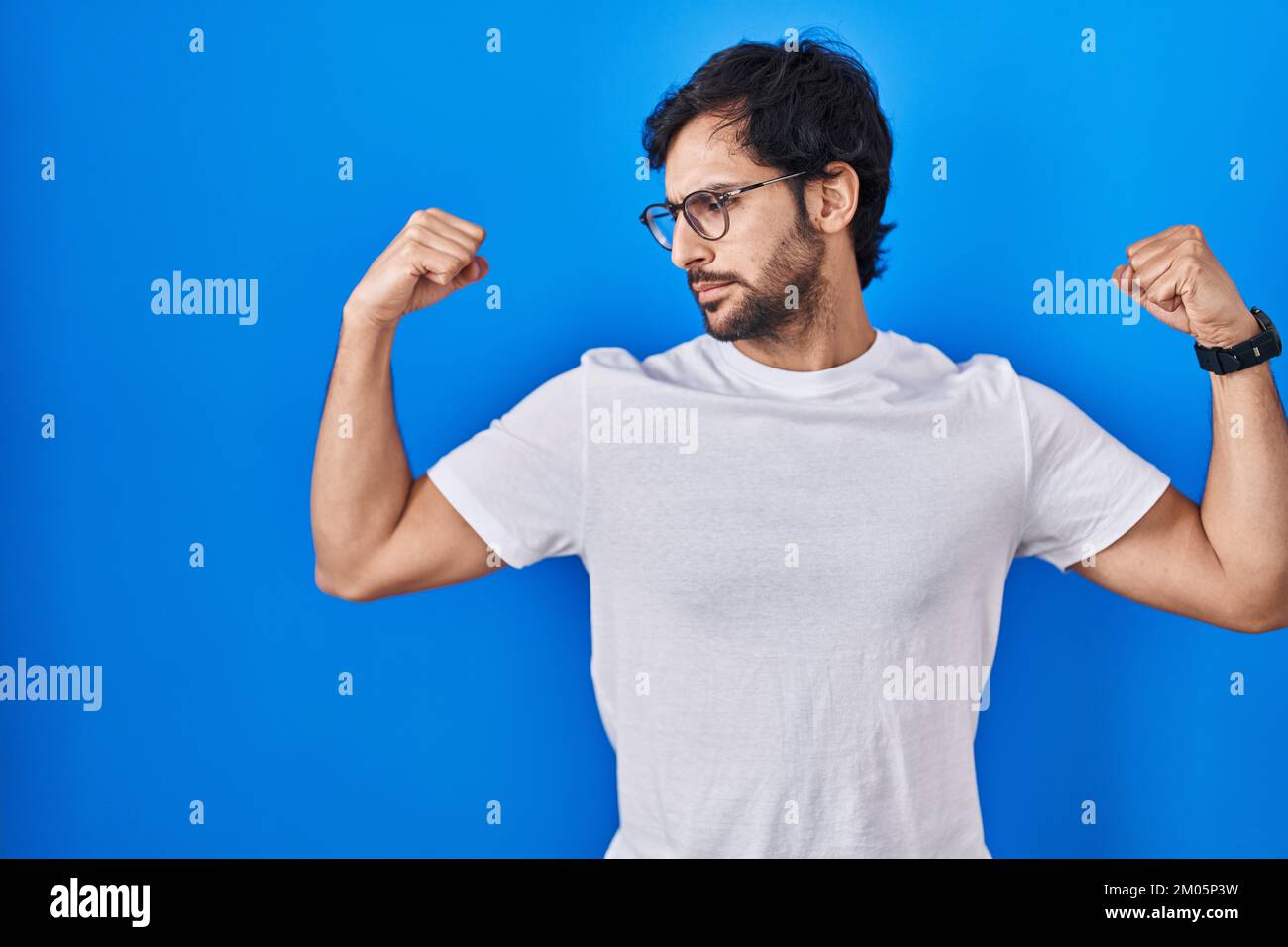 Handsome latin man standing over blue background showing arms muscles ...