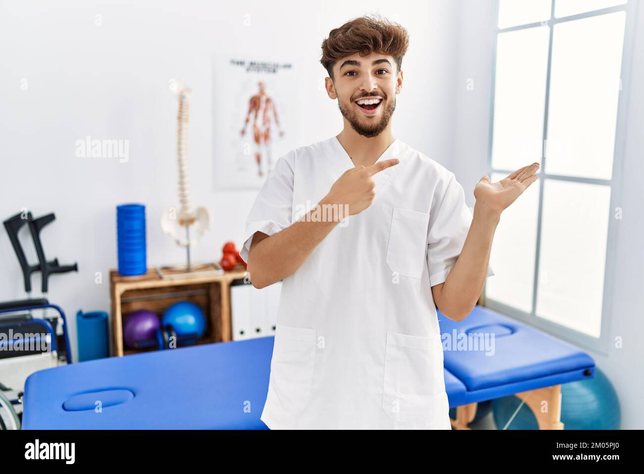 Young arab man working at pain recovery clinic amazed and smiling to ...