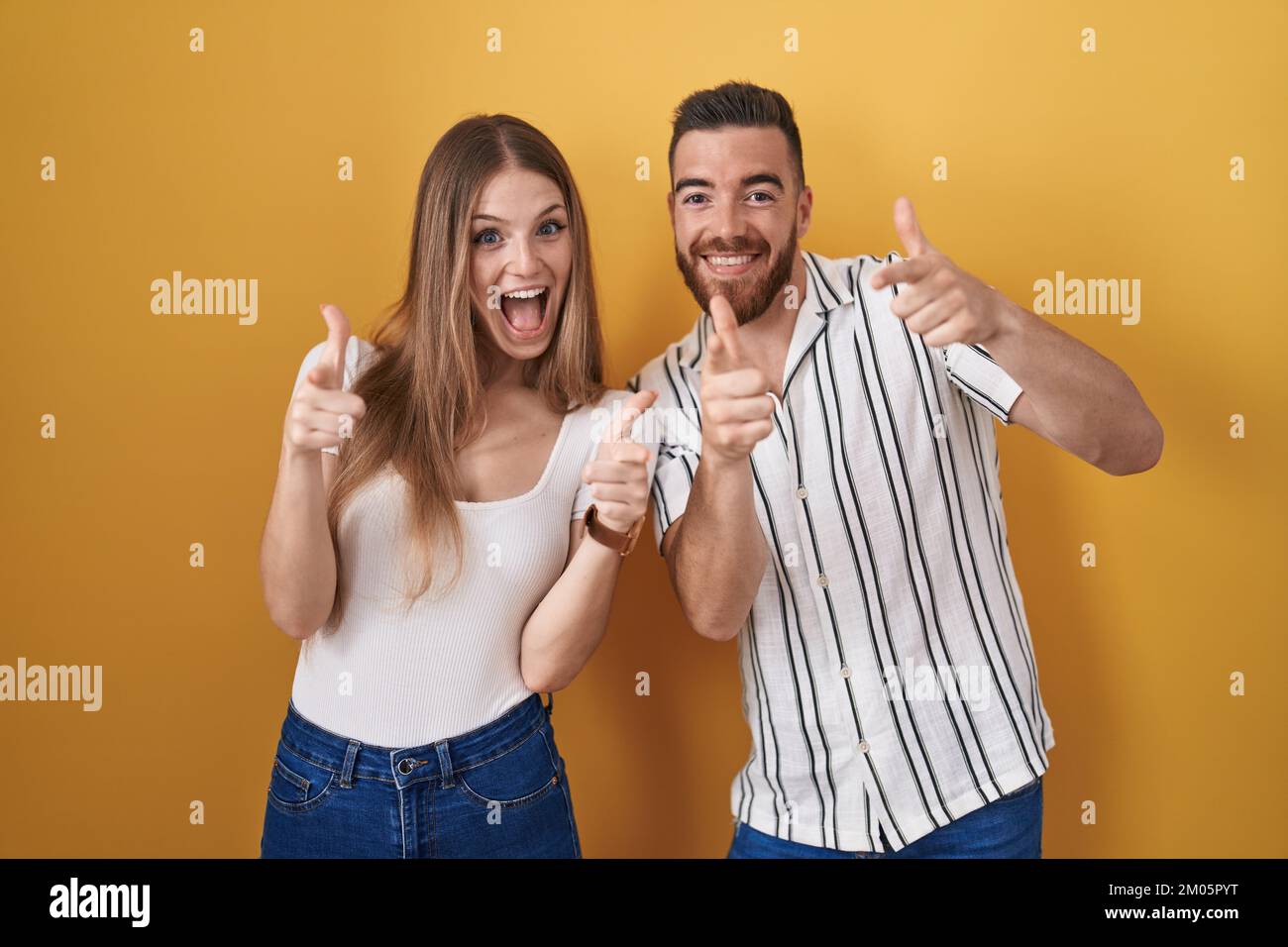 Young couple standing over yellow background pointing fingers to camera ...