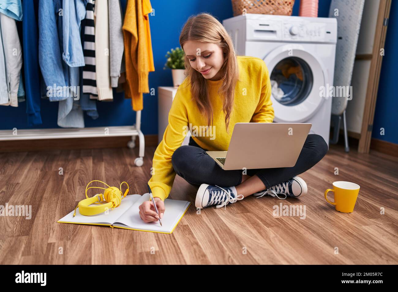Young blonde woman waiting for washing machine studying at laundry room ...
