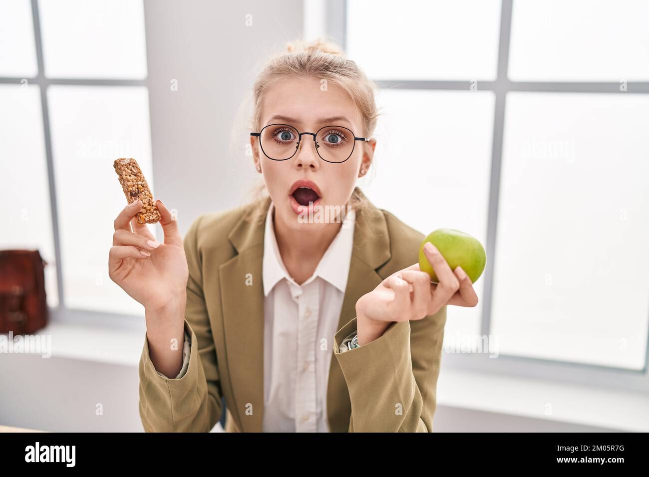 Young caucasian woman working at the office eating snack in shock face ...