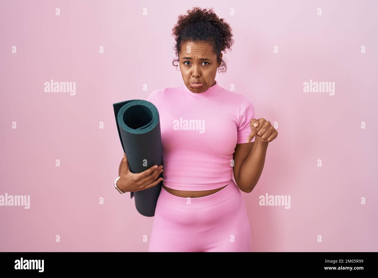 Young hispanic woman with curly hair holding yoga mat over pink ...