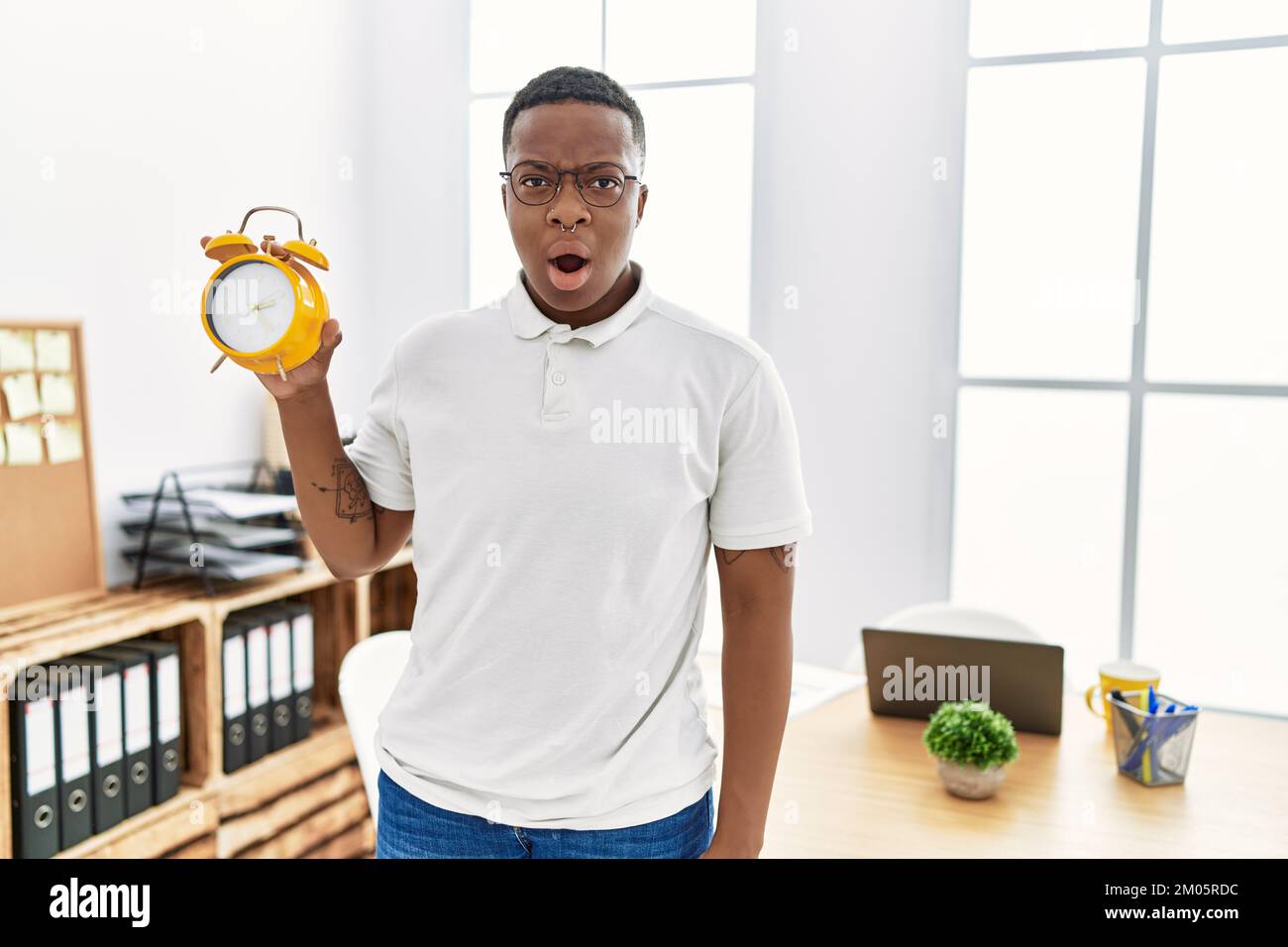 Young african man holding alarm clock at the office scared and amazed ...