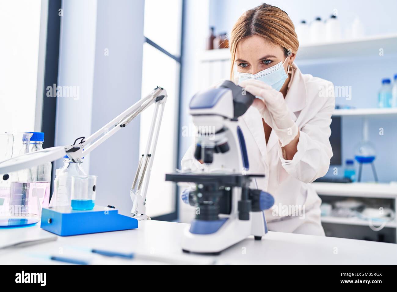 Young blonde woman scientist wearing medical mask using microscope at ...