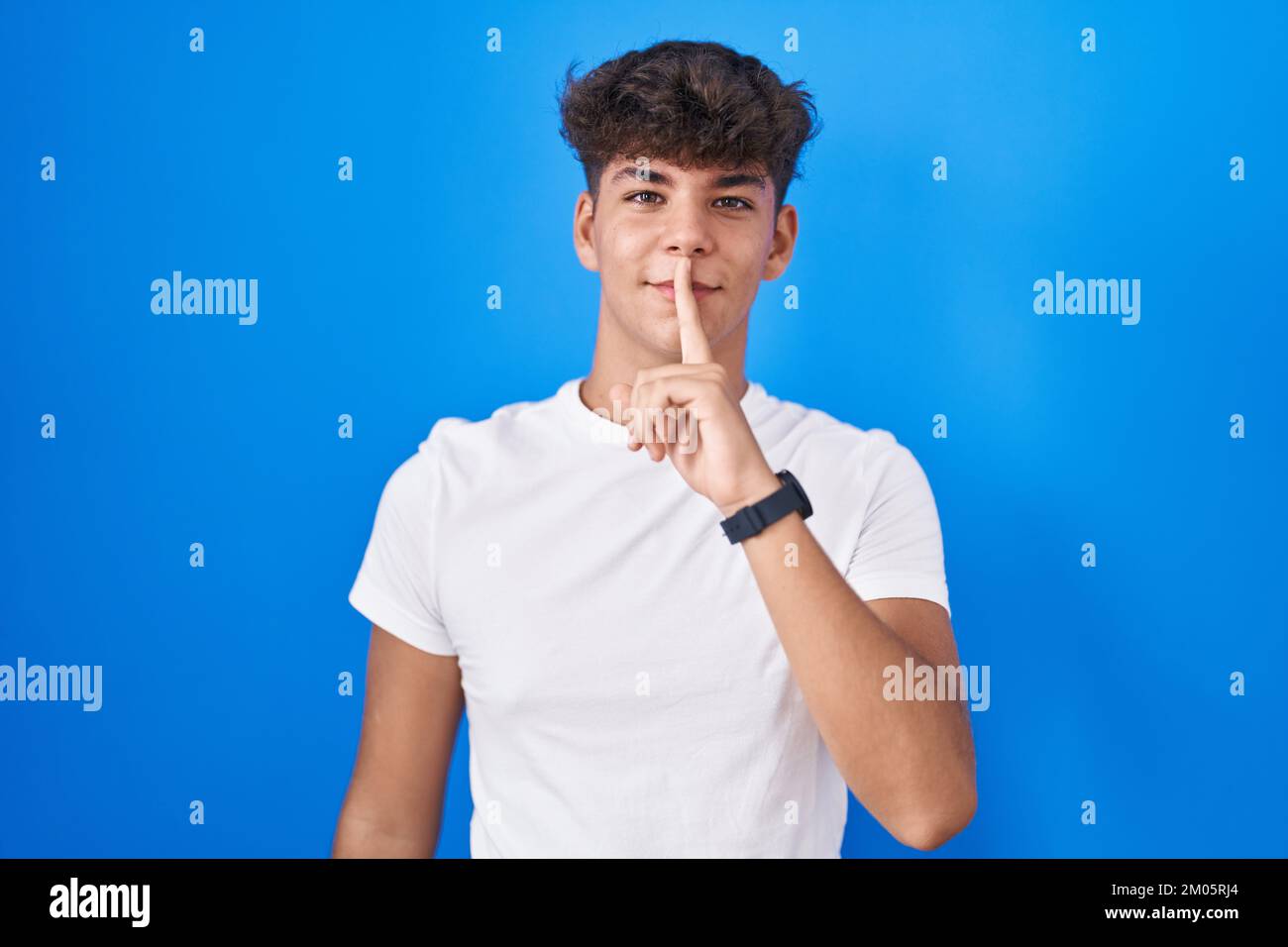 Hispanic teenager standing over blue background asking to be quiet with ...