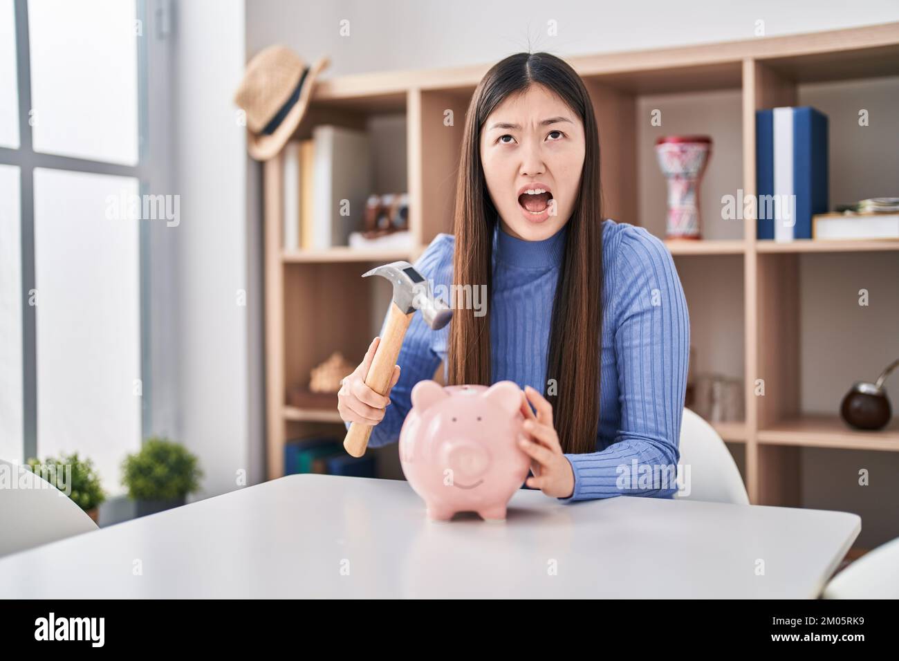 Chinese young woman holding hammer and piggy bank angry and mad ...