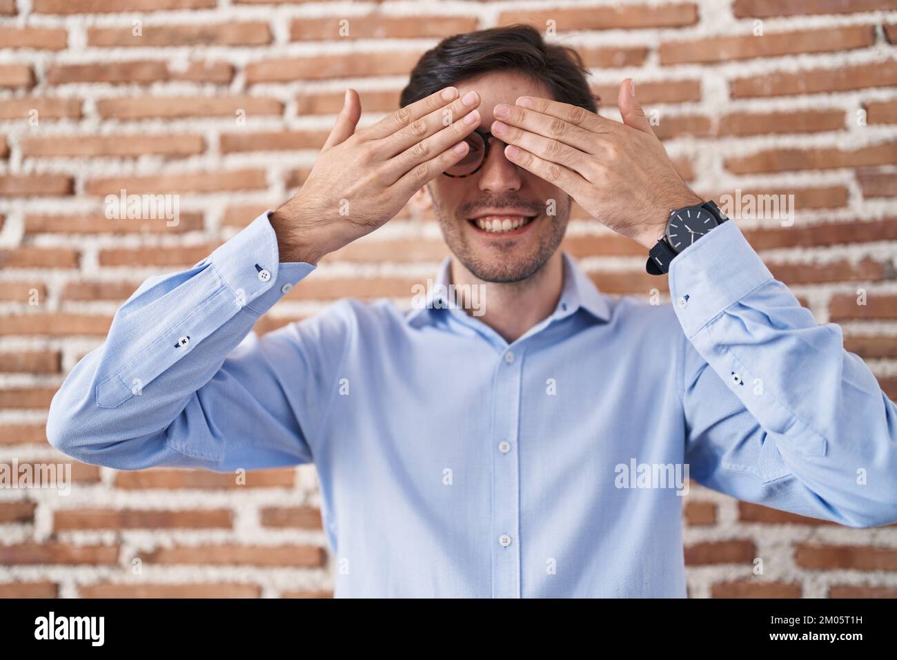 Young hispanic man standing over brick wall background covering eyes ...