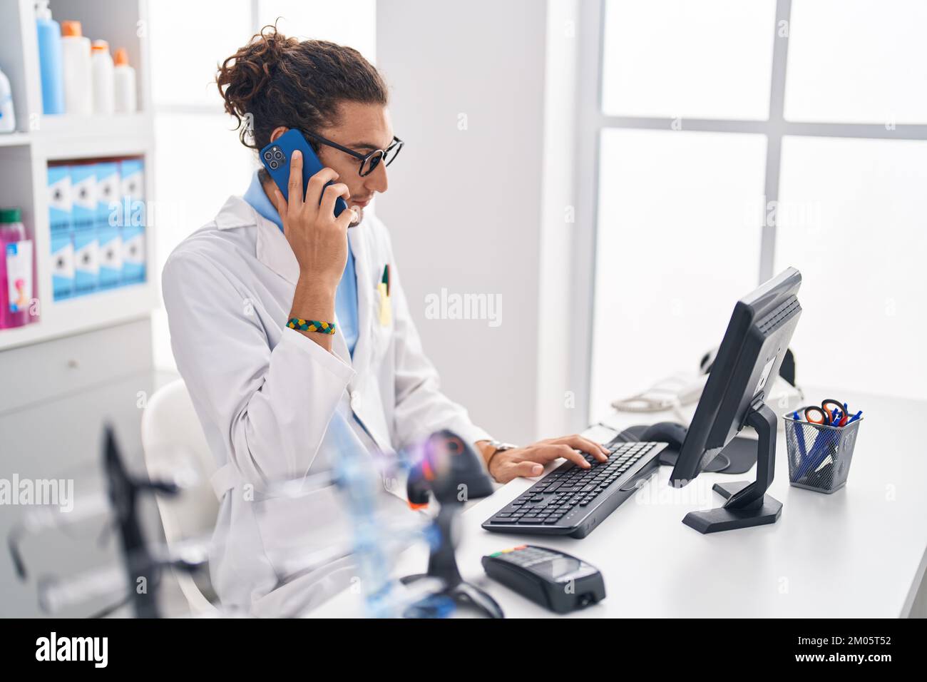 Young hispanic man pharmacist talking on smartphone using computer at ...