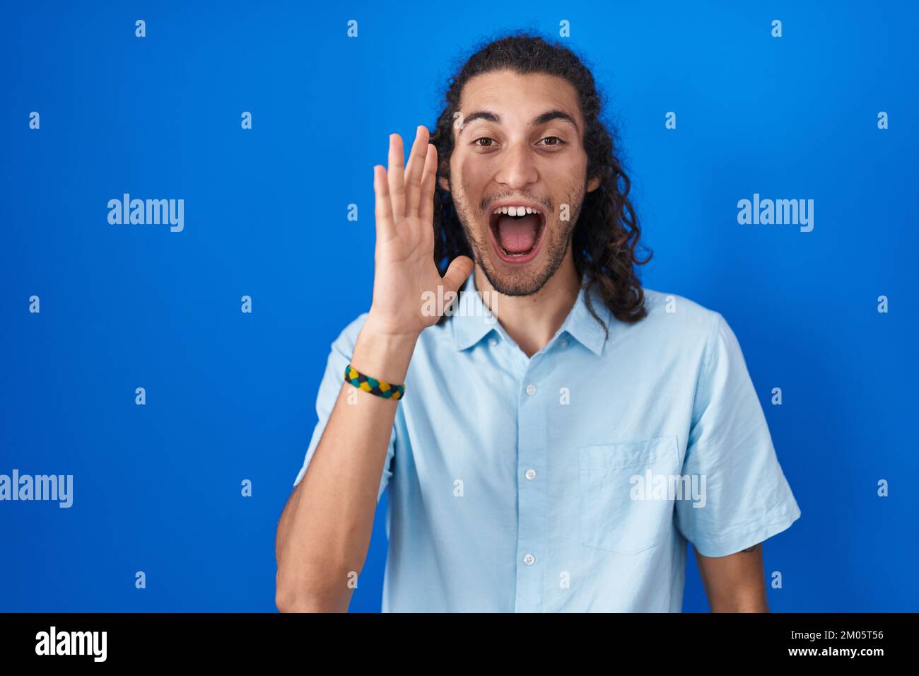 Young hispanic man standing over blue background shouting and screaming ...