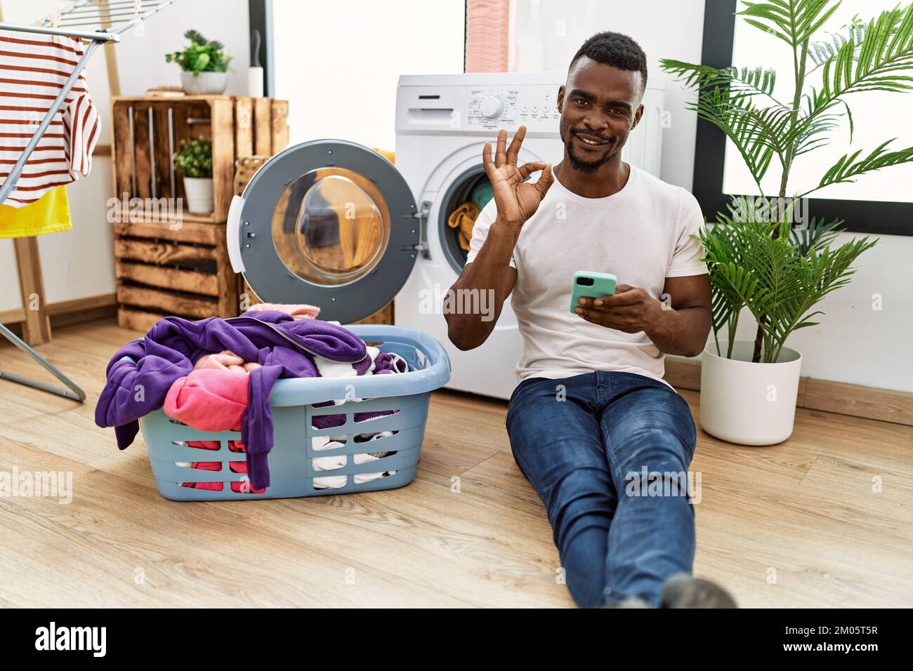Young african man doing laundry using smartphone doing ok sign with ...