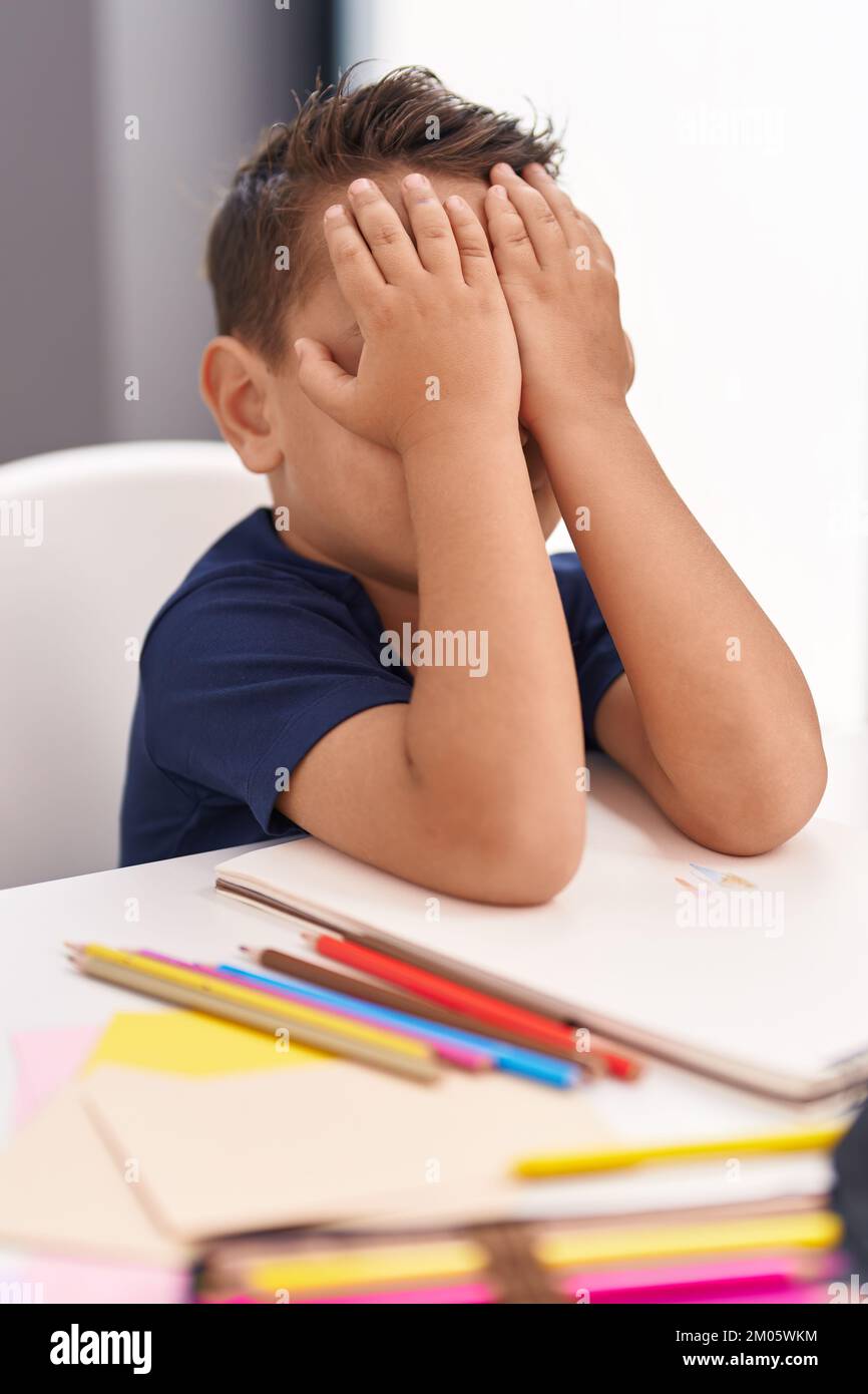 Adorable hispanic toddler student sitting on table crying at classroom ...