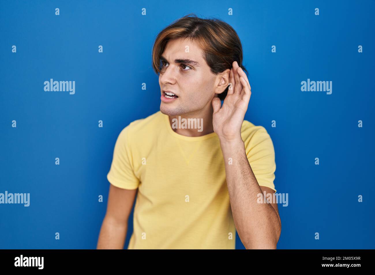 Young man standing over blue background smiling with hand over ear ...