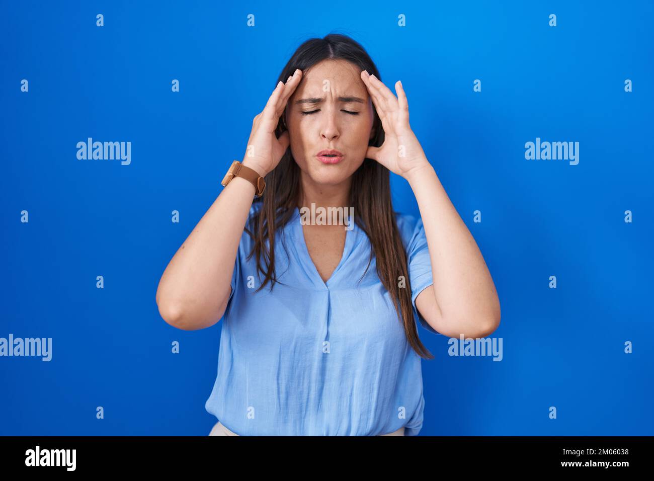 Young brunette woman standing over blue background with hand on head ...