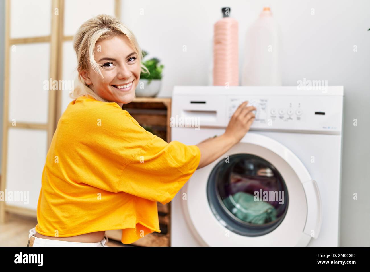Young blonde girl doing laundry putting clothes into washing machine at ...