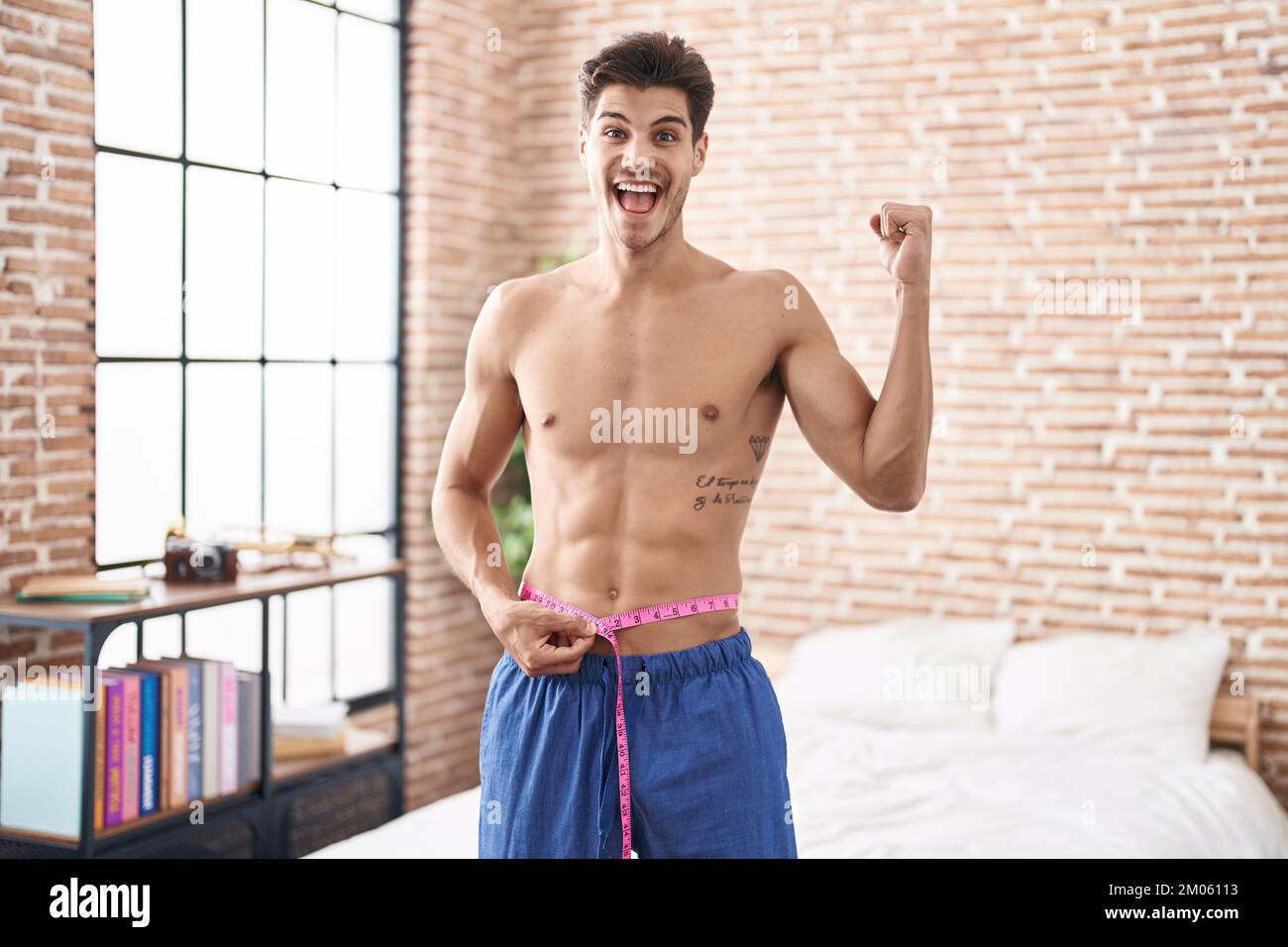 Young hispanic man using tape measure measuring waist screaming proud ...