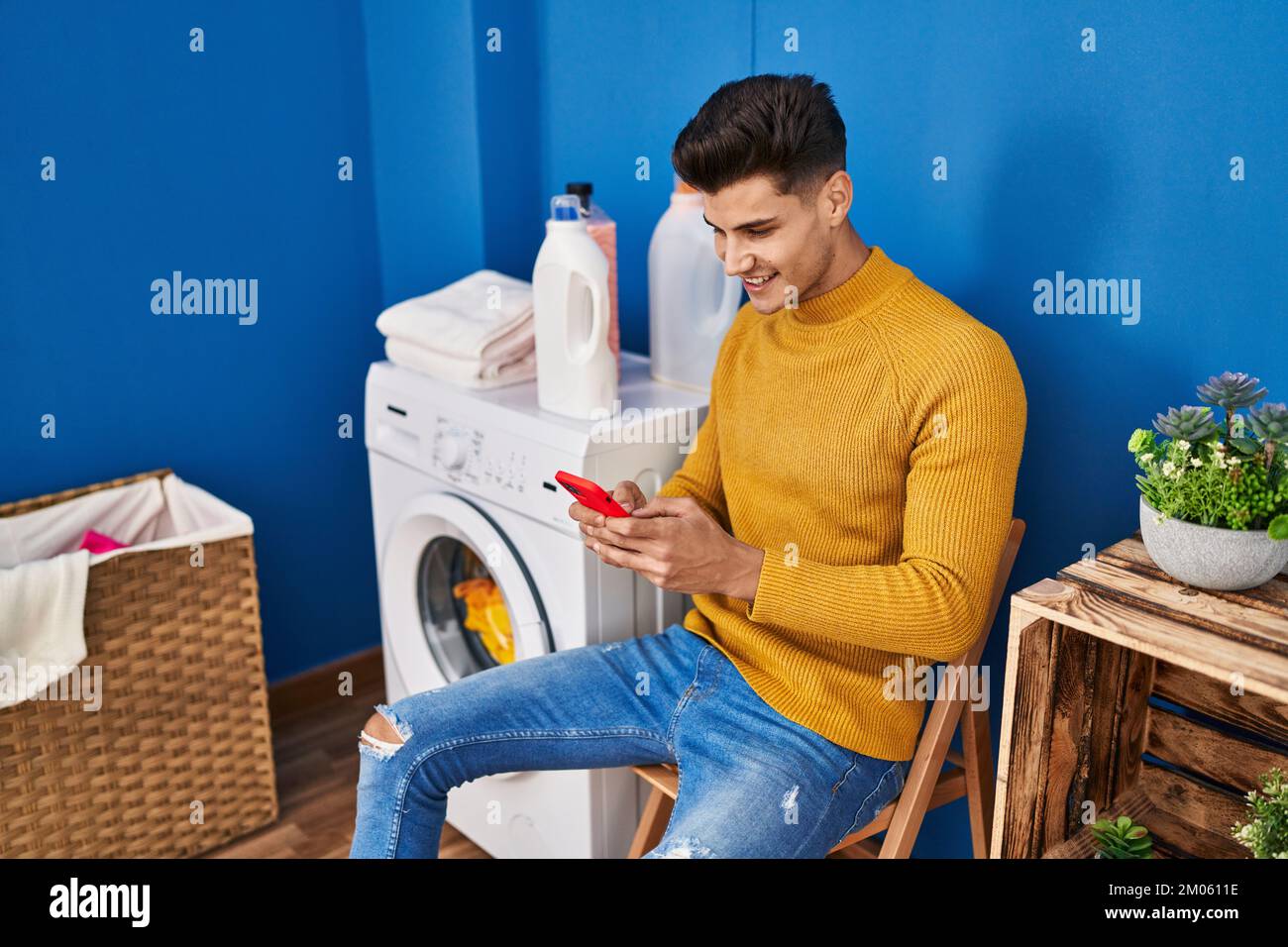 Young hispanic man using smartphone waiting for washing machine at ...