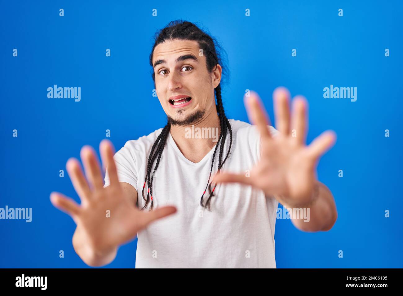 Hispanic man with long hair standing over blue background afraid and ...