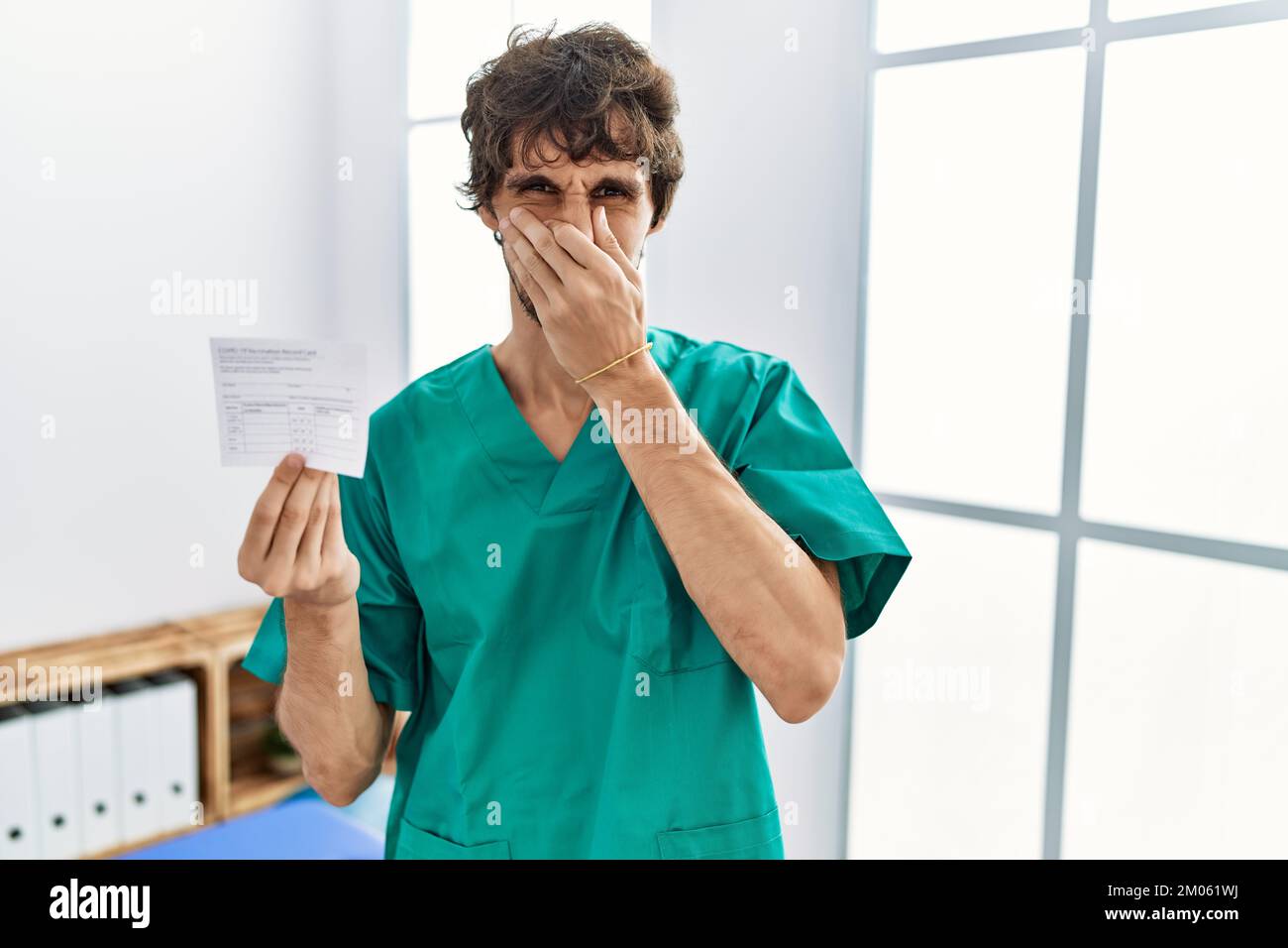 Young hispanic doctor man holding covid record card smelling something ...