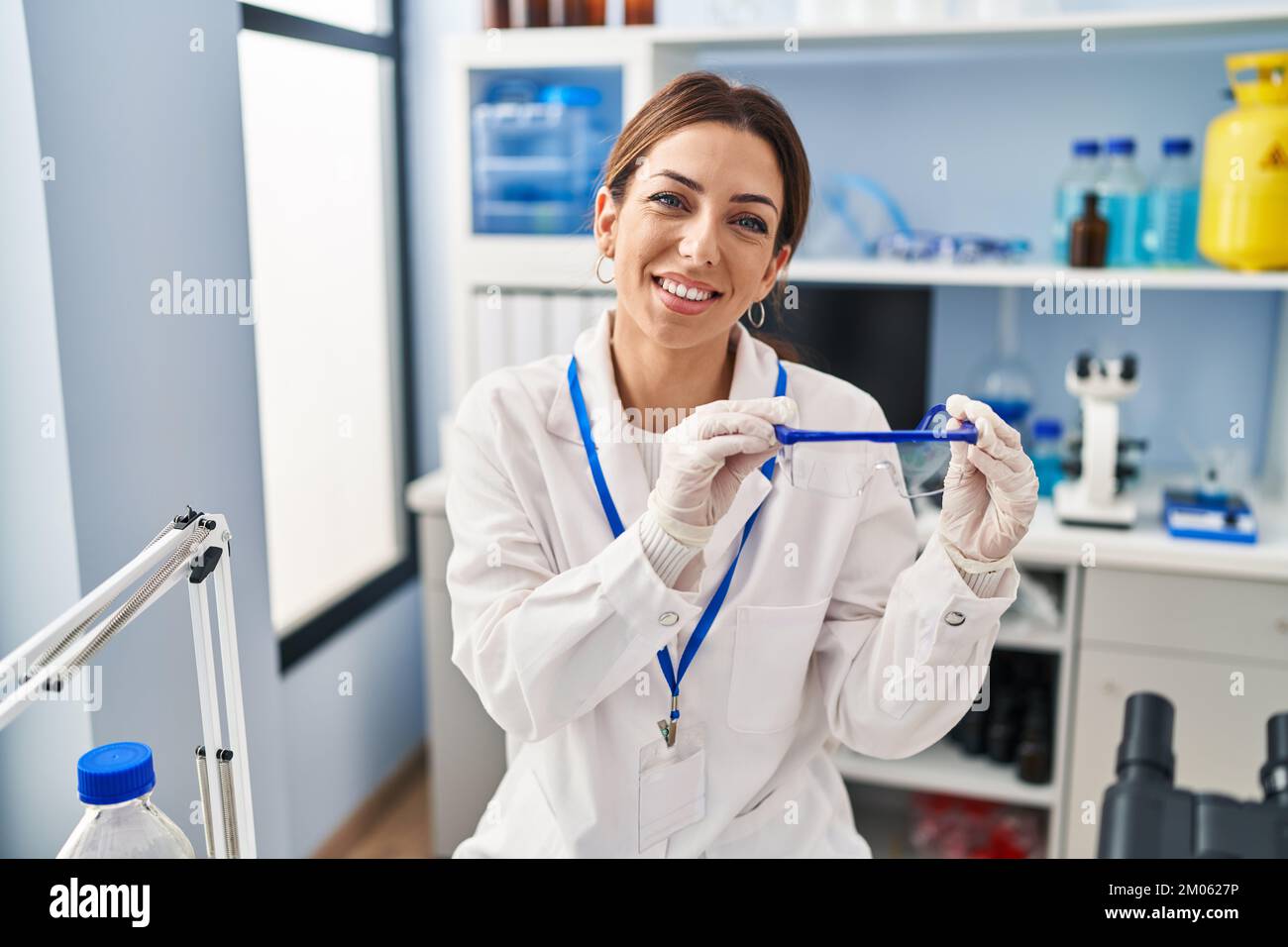 Young brunette woman working at scientist laboratory wearing safety glasses smiling with a happy ...