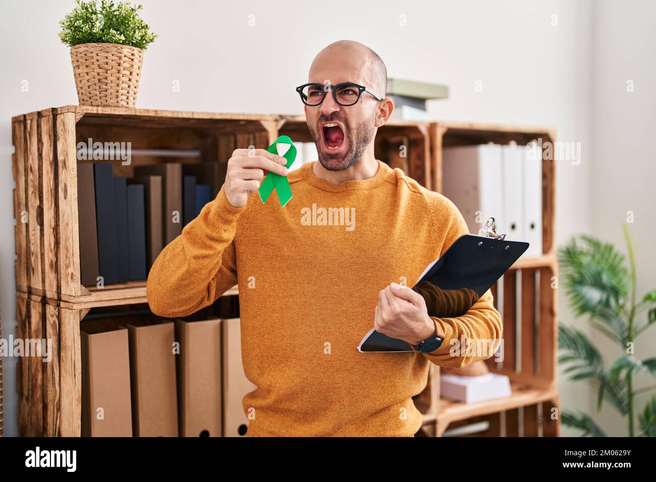 Young bald man with beard holding support green ribbon angry and mad ...