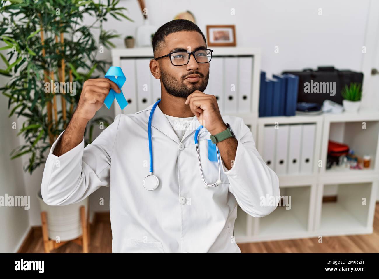 Young indian doctor holding blue ribbon serious face thinking about ...