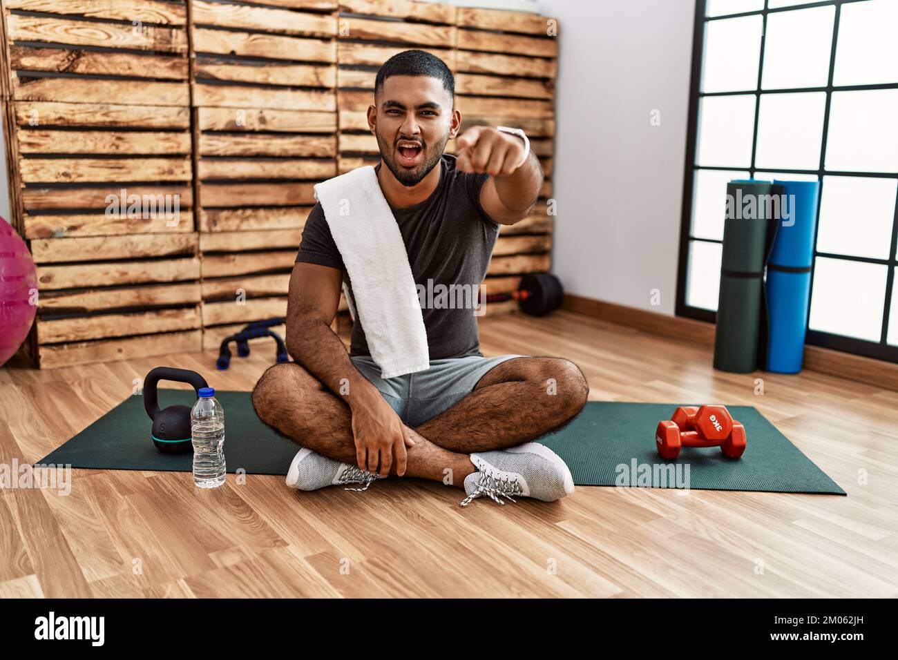 Young indian man sitting on training mat at the gym pointing displeased ...