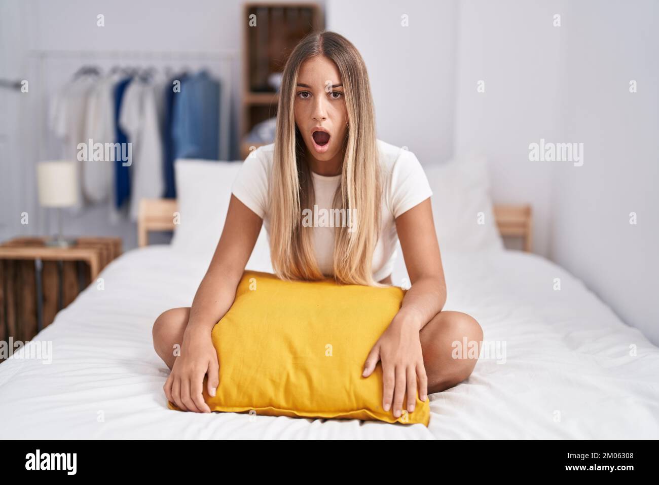 Young blonde woman sitting on the bed with pillow at home afraid and ...