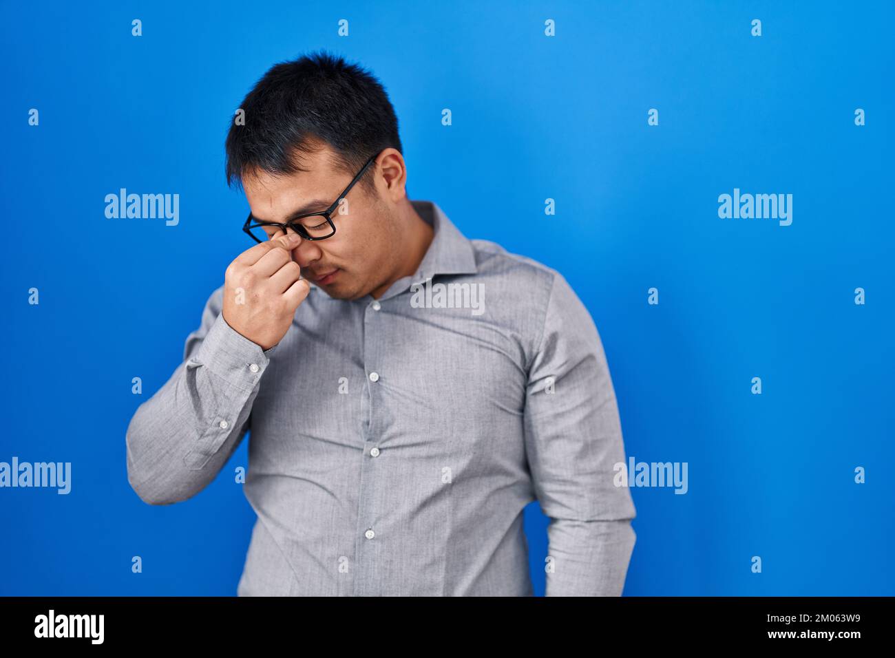 Young chinese man standing over blue background tired rubbing nose and ...